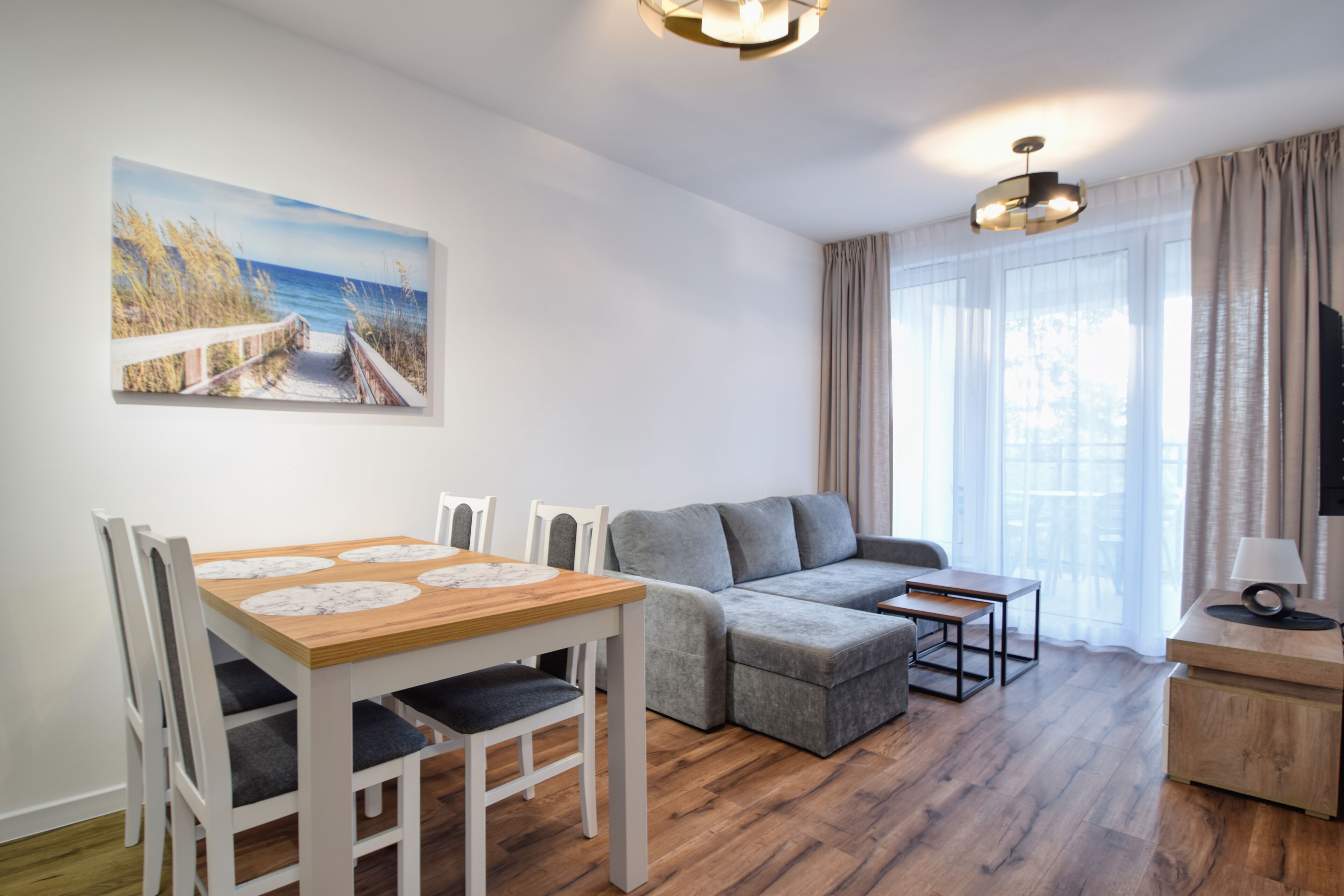 A modern living room with a wooden dining table, grey sofa, and a painting of a beach scene.