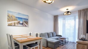 Uma sala de estar moderna com uma mesa de jantar de madeira, sofá cinza e um quadro com uma cena de praia.