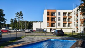 Piscina com deck de madeira, cercada por um estacionamento com vários carros. Ao fundo, um edifício de vários andares com varandas.