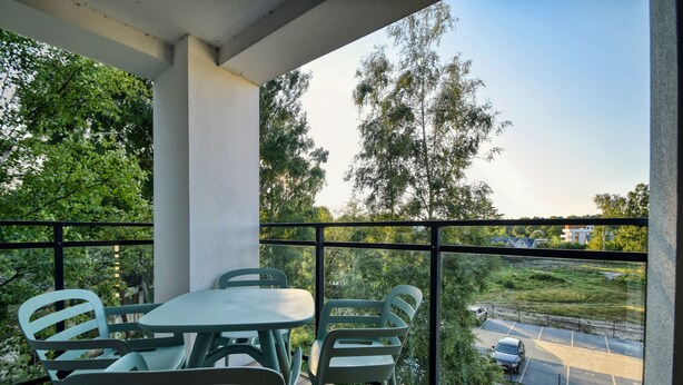 Varanda com mesa e cadeiras, com vista para um estacionamento e árvores.