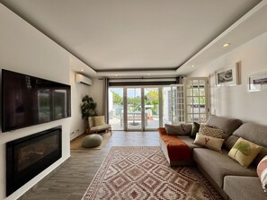 Uma sala de estar moderna com televisão de tela plana, lareira, sofá com almofadas, tapete estampado e vista para área externa por meio de portas de vidro.