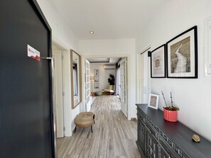 Um corredor com uma porta escura, uma cadeira de vime, uma cama com cabeceira e um aparador com quadros e uma planta em vaso.