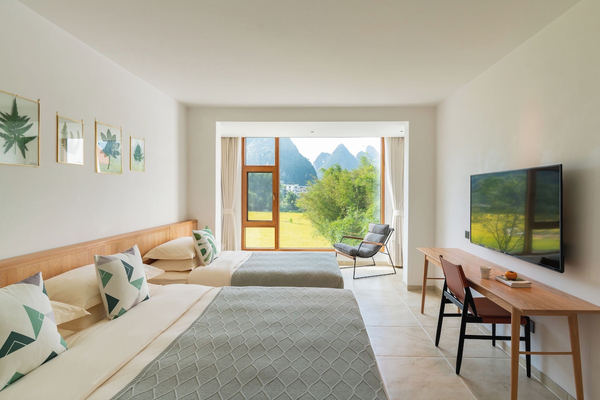Quarto de hotel com duas camas, uma escrivaninha, uma cadeira, televisão e uma janela grande com vista para as montanhas.