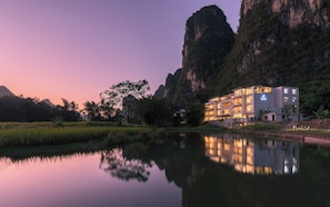 Um hotel moderno com vários andares, localizado à beira de águas calmas, rodeado por vegetação exuberante e montanhas.