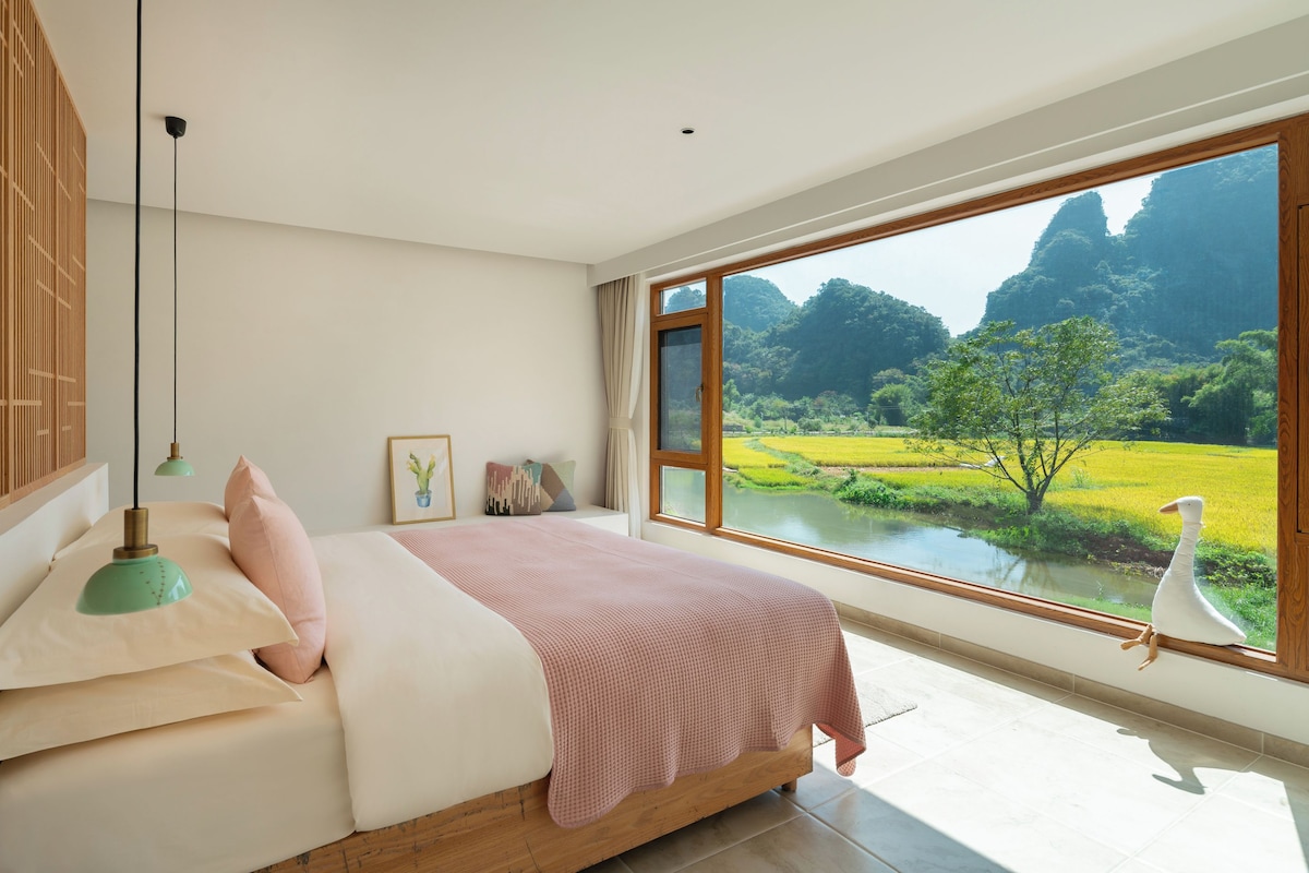 Quarto com uma grande janela, uma cama, mesa de cabeceira e vista para a paisagem.