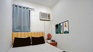 A bedroom with a bed, a bedside table with a phone and a vase of flowers, a wall-mounted air conditioner, and a window with a curtain.