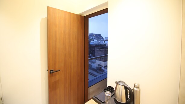 Quarto com porta de madeira, janela com vista para prédios, chaleira e garrafa de água.