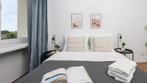 Quarto de hotel com uma cama, duas mesas de cabeceira, uma janela com cortinas e dois quadros na parede.