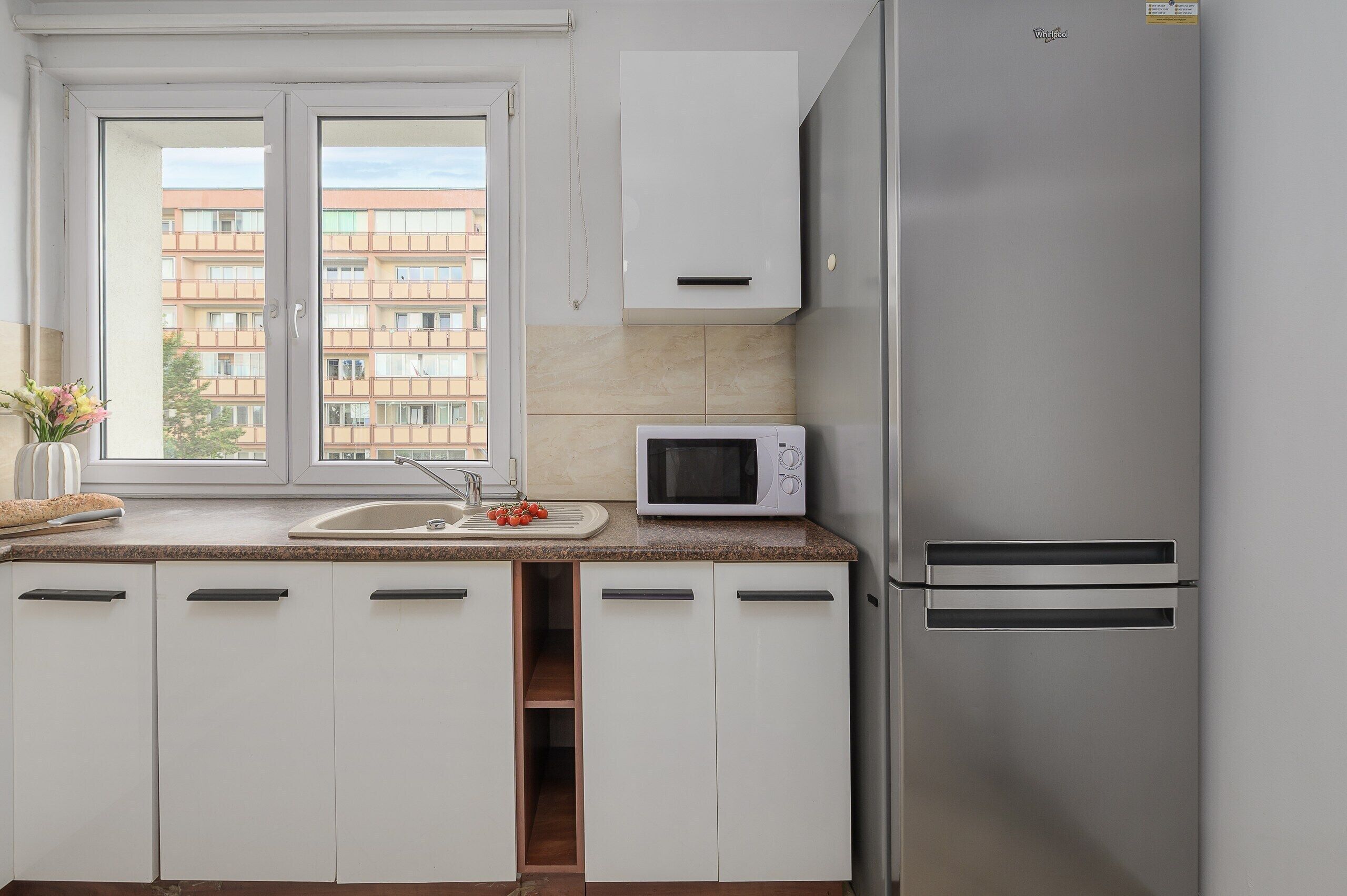 Uma cozinha moderna com armários brancos, geladeira de aço inoxidável, micro-ondas e janela com vista para um edifício.