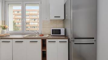 Uma cozinha moderna com armários brancos, geladeira de aço inoxidável, micro-ondas e janela com vista para um edifício.