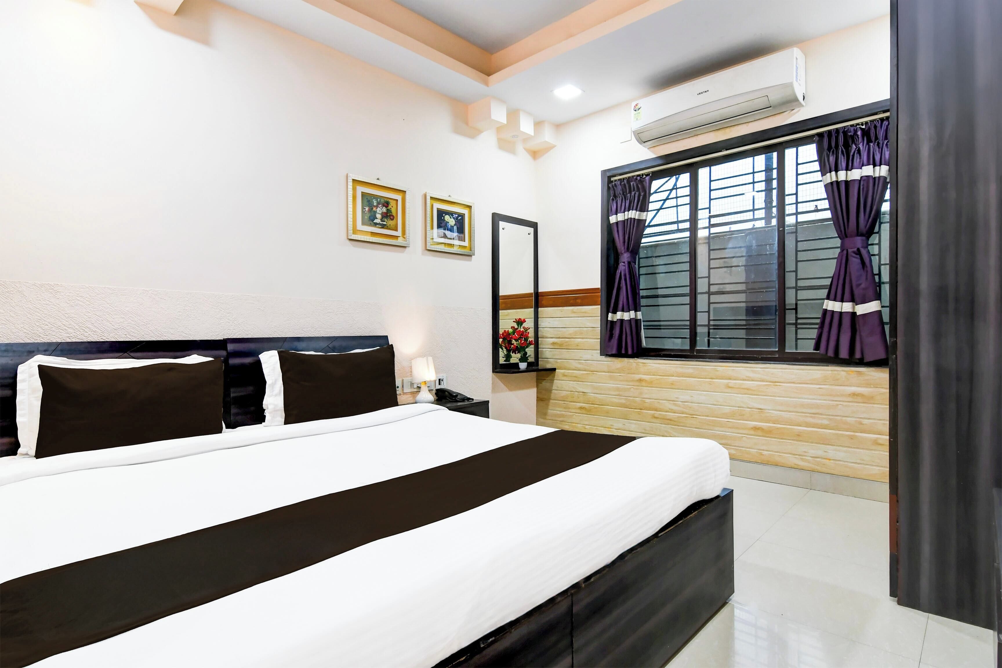 Quarto de hotel com uma cama grande, uma mesa de cabeceira com abajur, um espelho e uma janela com cortinas.