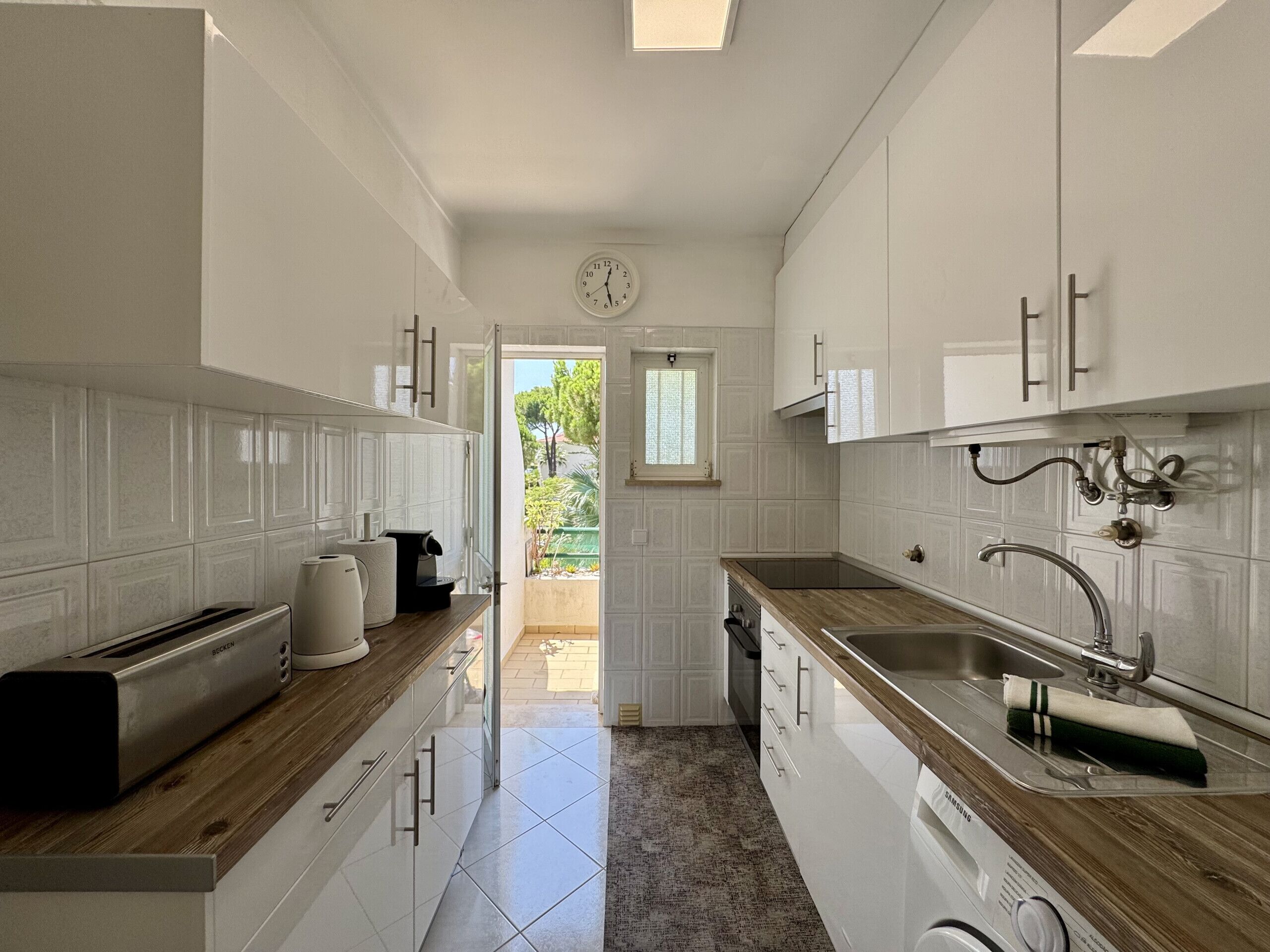 Cozinha moderna com armários brancos, forno embutido, pia e janela com vista para um jardim.