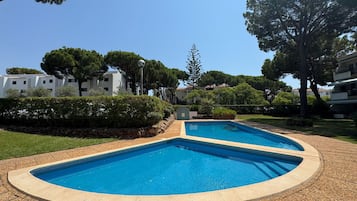 Uma área de piscina com duas piscinas, rodeada por um caminho pavimentado, árvores e edifícios ao fundo.