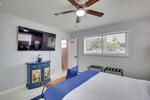 Quarto com cama, televisão montada na parede, ventilador de teto e janela com persianas.