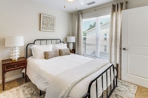 Quarto com cama, duas mesas de cabeceira, luminária na parede, cortina estampada e uma porta.