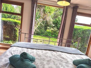 Quarto com cama, vista para um jardim através de grandes janelas e guarda-corrimão de metal.