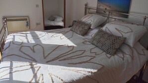 Quarto com uma cama, um quadro de uma praia e uma porta que dá para outro cômodo.