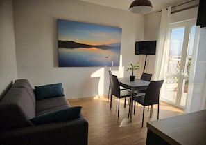 Uma sala de estar com sofá, mesa de jantar com cadeiras, um quadro na parede e TV fixada na parede.