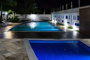 Piscina com deck revestido, espreguiçadeiras e um edifício com várias janelas ao fundo.