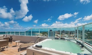 Terraço no telhado com hidromassagem, móveis de vime e guarda-corpo de vidro, com vista para a cidade e o oceano.