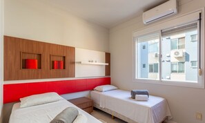 Quarto de hotel com cama, uma mesinha, um banco, janela com vista para o exterior e ar-condicionado fixado na parede.