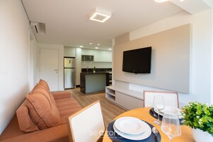 Uma sala de estar moderna com um sofá, uma mesa de jantar posta com pratos e copos, e uma televisão na parede.