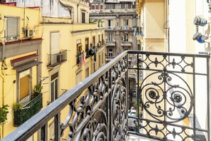 Varanda com grades de metal ornamentadas, dando para uma rua estreita com edifícios.