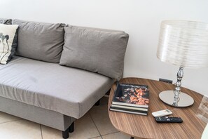 Uma sala de estar moderna com um sofá cinzento, uma mesa de madeira redonda pequena com uma luminária, livros e um controle remoto.
