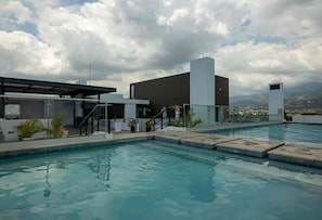 Piscina com um edifício moderno ao fundo, céu claro e montanhas ao longe.