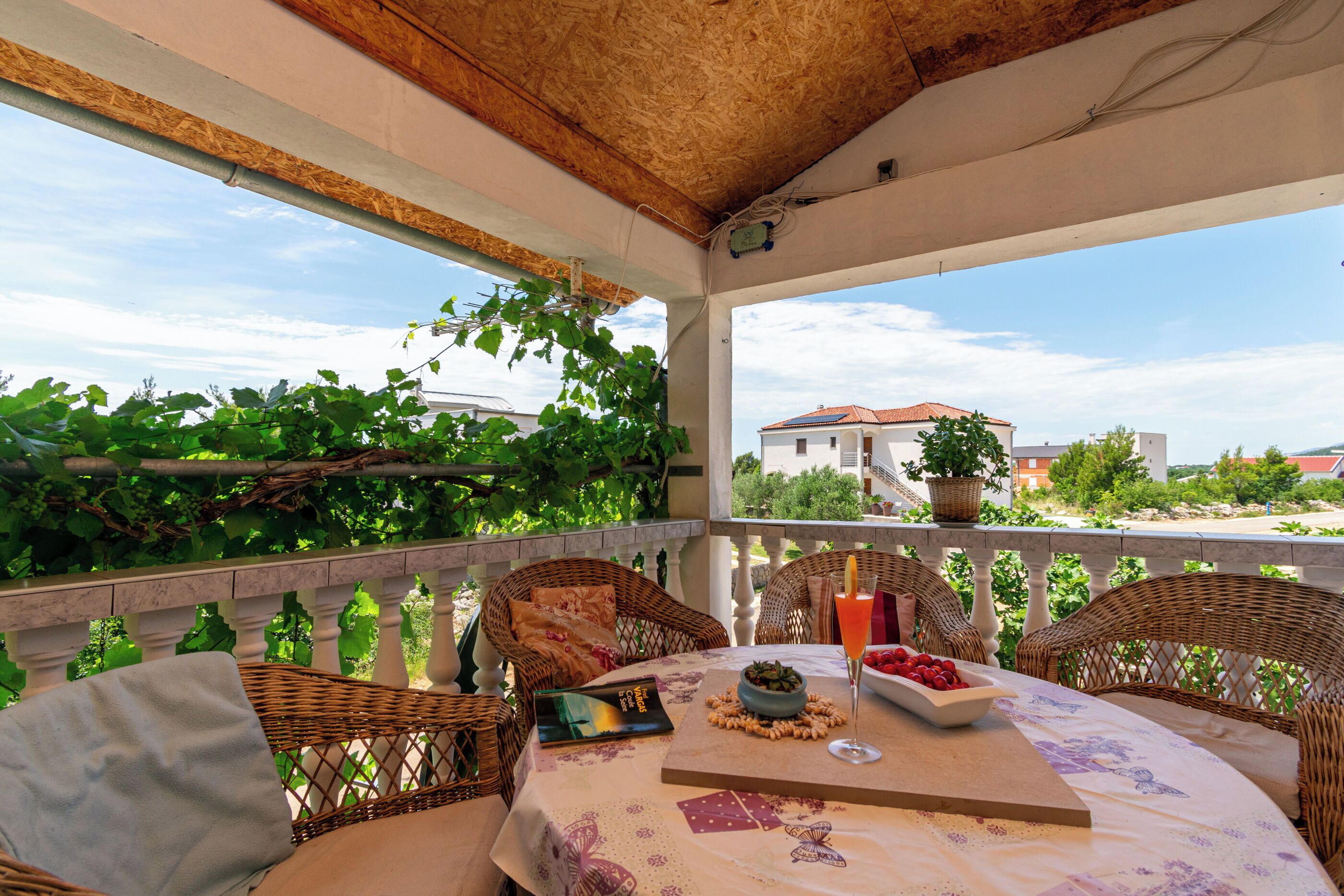 Uma área de estar coberta ao ar livre com móveis de vime, uma mesa com uma cesta de frutas e uma bebida, e vista para edifícios ao fundo.