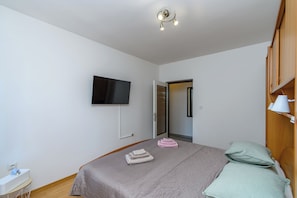 Quarto com cama, televisão na parede, mesa de cabeceira e porta que dá para outro cômodo.