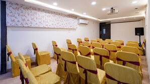 Uma sala de conferências com fileiras de cadeiras cobertas com tecido amarelo e fitas vermelhas, paredes brancas com papel de parede floral, um aparelho de ar condicionado e um projetor fixado ao teto.