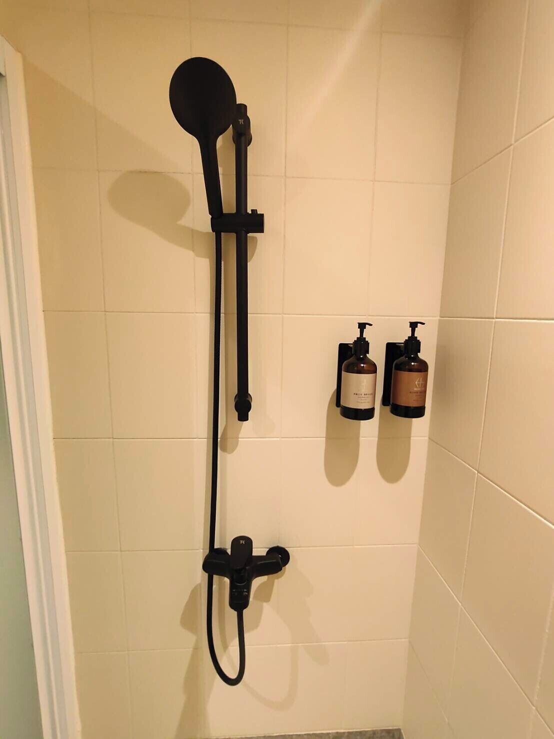 Um chuveiro com um chuveiro manual e um massageador manual. Há duas garrafas de shampoo e condicionador na parede.