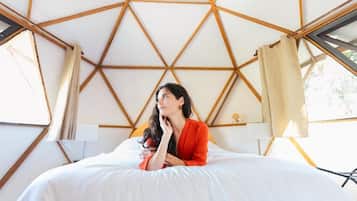 Uma pessoa sentada em uma cama dentro de uma estrutura parecida com uma tenda, com uma estrutura de madeira geométrica.