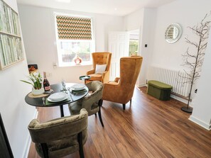 Uma área de refeições com uma mesa posta para dois, uma cadeira, uma pequena mesa lateral e uma planta decorativa.