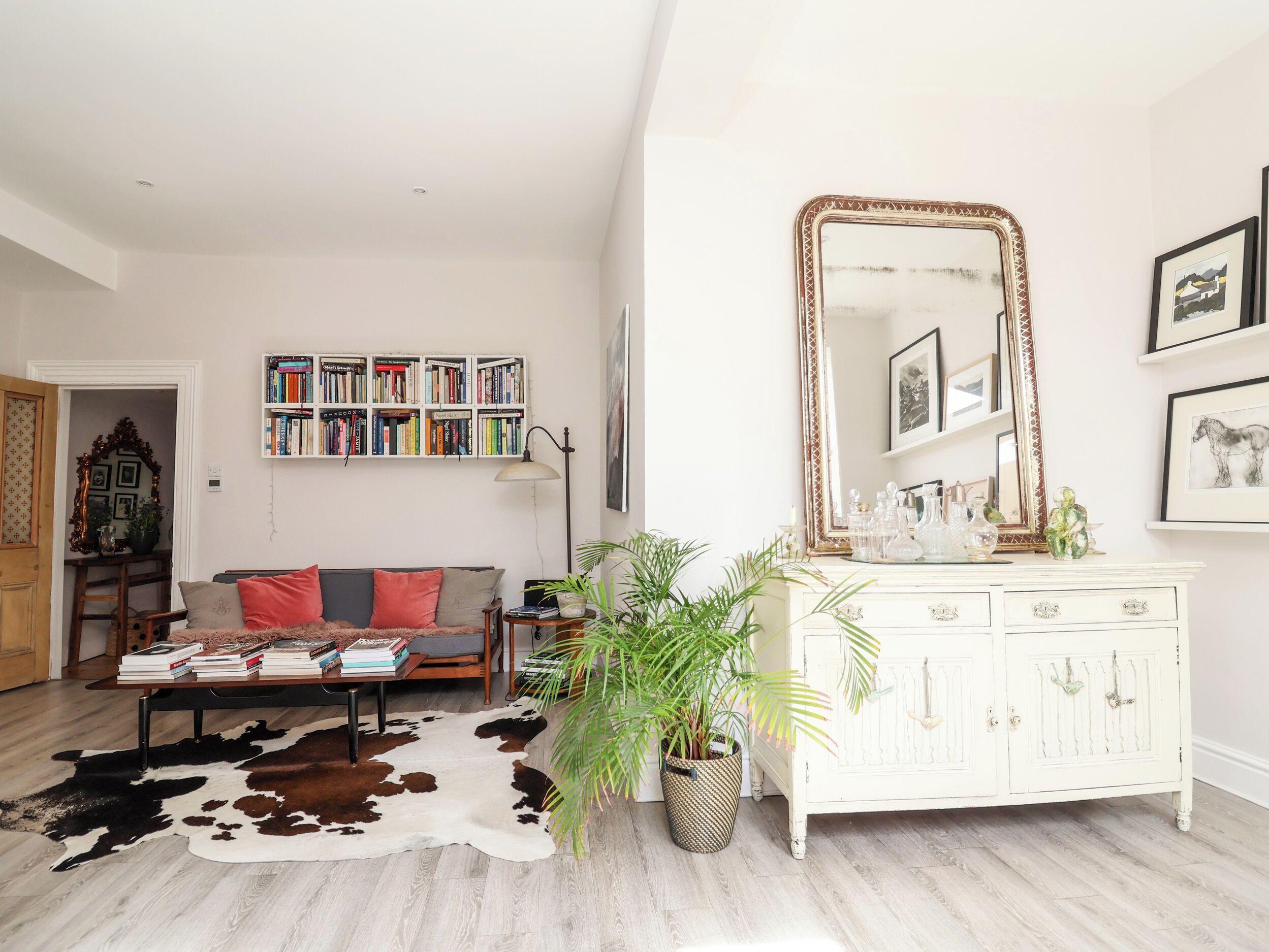 Uma sala de estar com um sofá branco, um tapete de couro de vaca, uma estante de livros, um armário branco e um espelho grande.