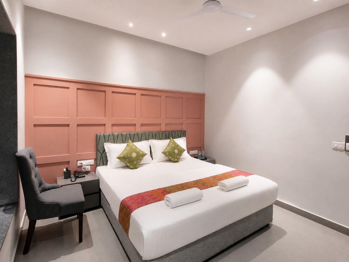 Quarto de hotel com cama, escrivaninha, cadeira, ventilador de teto e mesa de cabeceira.