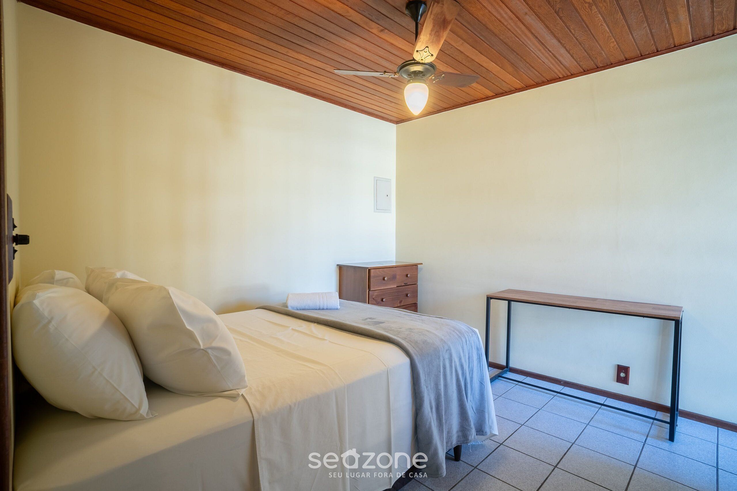Quarto com cama, mesa de cabeceira, cômoda, ventilador de teto e janela.