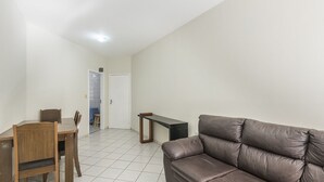 Uma sala de estar com sofá de couro marrom, mesa de jantar de madeira com cadeiras, uma pequena mesa lateral preta e uma porta branca que dá para outro cômodo.