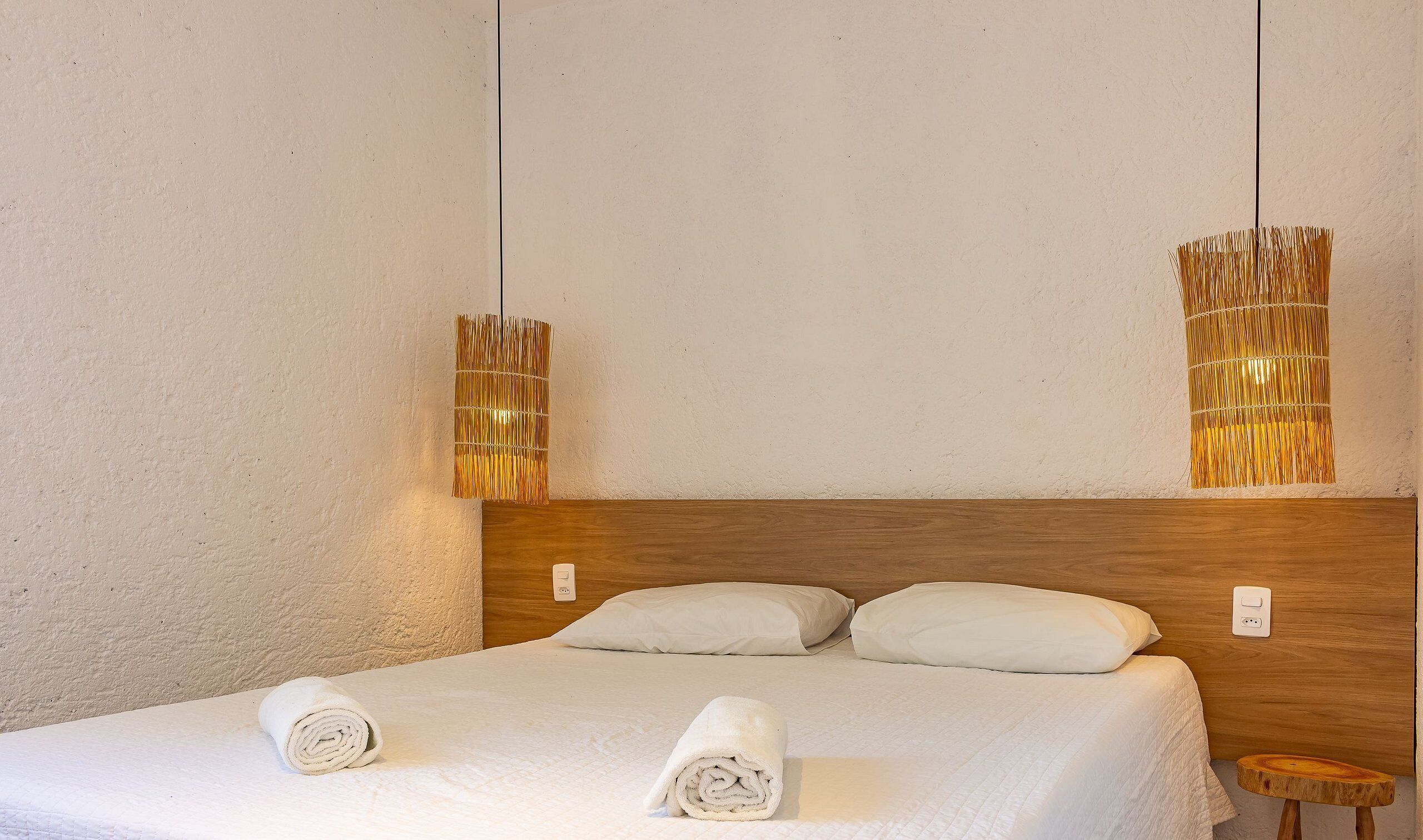 Cama bem arrumada com duas toalhas enroladas, cabeceira de madeira e duas luminárias fixadas na parede.