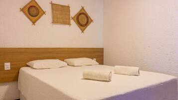 Cama bem arrumada com lençóis brancos e toalhas dobradas, cabeceira de madeira e três tapeçarias de parede.