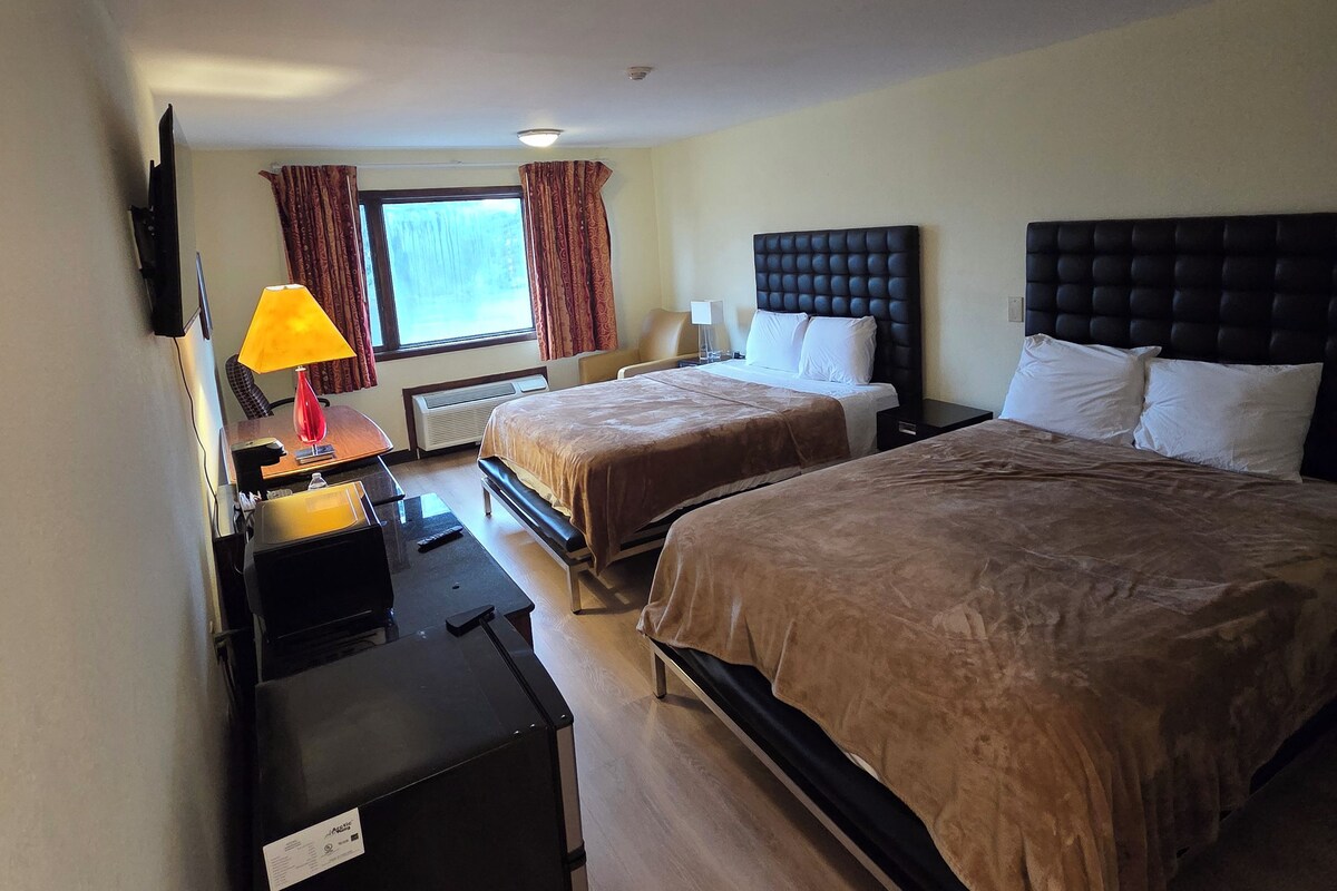 Quarto de hotel com duas camas, uma escrivaninha, uma cadeira, uma luminária e uma janela com cortinas.