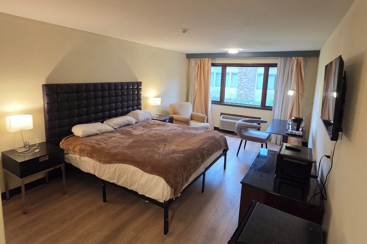 Quarto de hotel com uma cama grande, mesas de cabeceira, escrivaninha, cadeira, televisão e janela com cortinas.