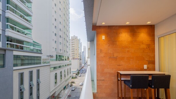Uma varanda com mesa e cadeiras, com vista para uma rua da cidade com prédios e carros estacionados.