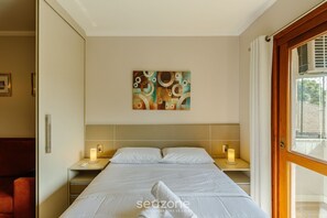 Quarto de hotel com cama, luminárias de cabeceira, um quadro na parede e vista através de uma porta de vidro.
