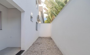 Um corredor exterior estreito, com paredes brancas, chão de cascalho e um pequeno degrau que dá para uma porta.