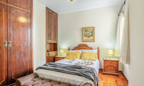 Um quarto com guarda-roupa de madeira, cama com almofadas amarelas, mesa de cabeceira com abajur e um quadro na parede.