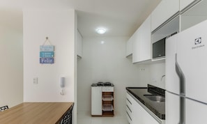 Cozinha compacta com armários brancos, bancada preta e geladeira de aço inoxidável. Há uma pequena ilha com pia e um telefone fixado na parede.