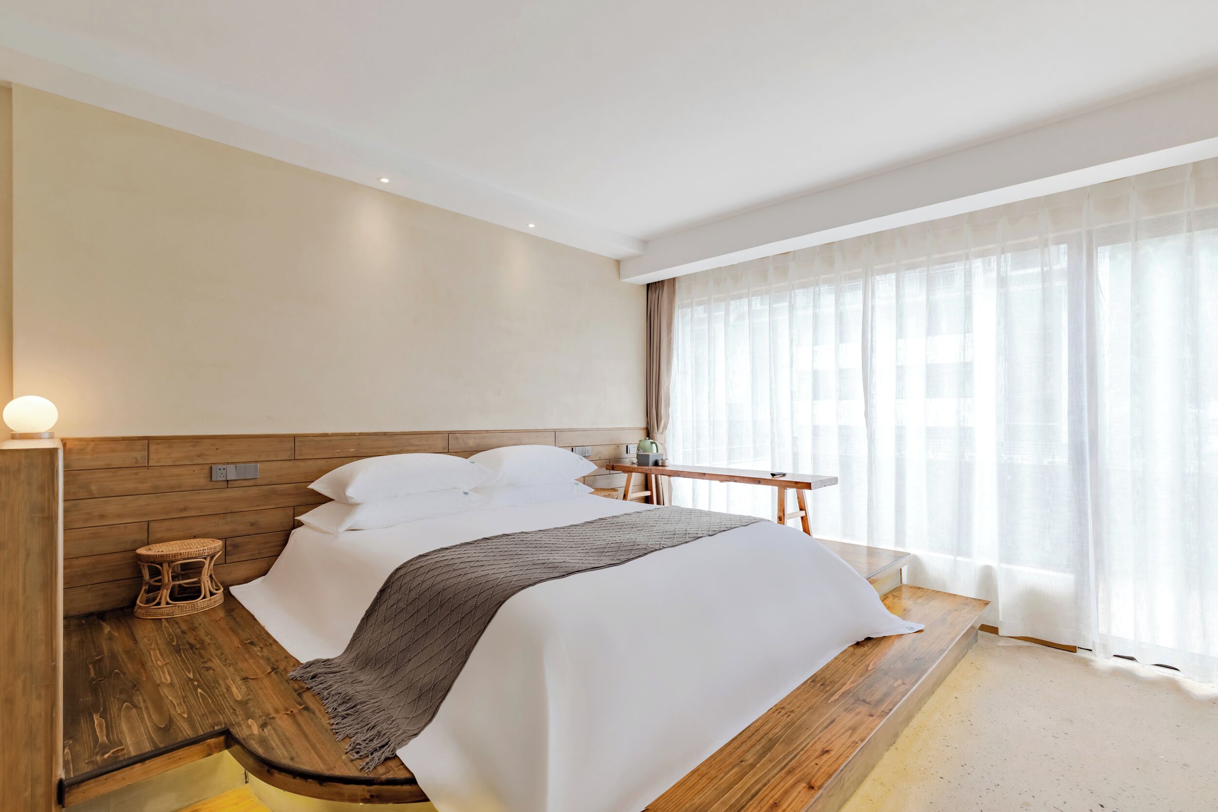 Quarto de hotel com cama, mesa de cabeceira, abajur e vista da cidade através de cortinas translúcidas.