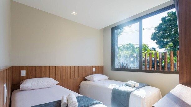 Quarto de hotel com duas camas, cabeceira de madeira, janela grande com vista para árvores e uma pequena escrivaninha com abajur.